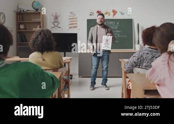 Tracking in of male Caucasian teacher holding image of frog anatomy, teaching high school ...