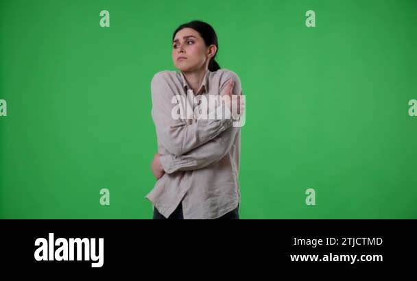 A Caucasian woman stands on a green screen background, hugging herself ...
