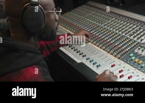 Young black man in casualwear and headphones sitting by sound mixer and ...