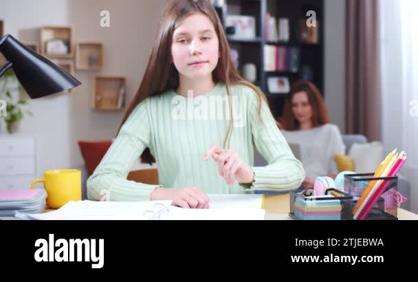Close up teenager school girl doing hometask, looking at camera and smiling. Daughter writing ...
