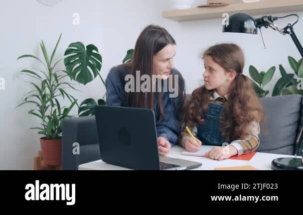 Mother watching kid writing in copybook at table with laptop, book ...