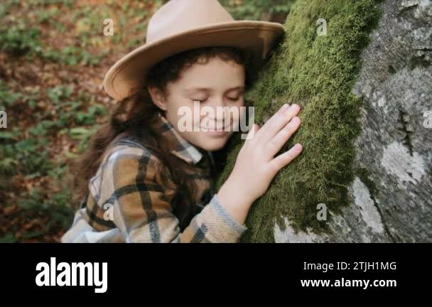 Happy cute little girl child in hat girl touch softly old tree bark ...