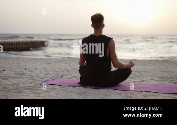 A young man is doing yoga and meditating on a sports mat while sitting ...