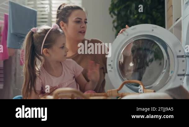 Happy mother and daughter put clothes in washing machine. Woman closed the washing machine door ...