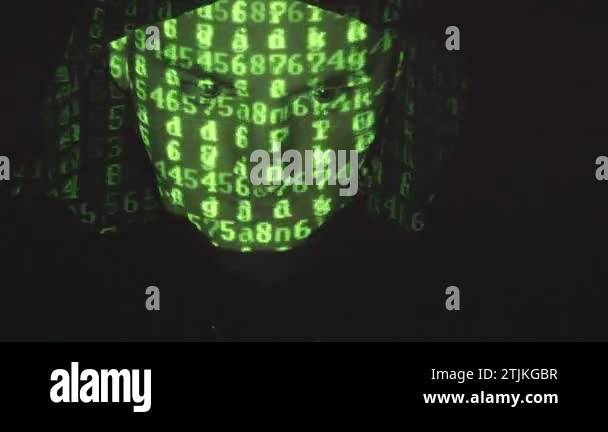 Face of male hacker in hood working on a computer while green code ...