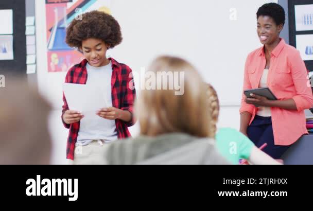 Diverse female teacher and happy schoolchildren at desks reciting in ...