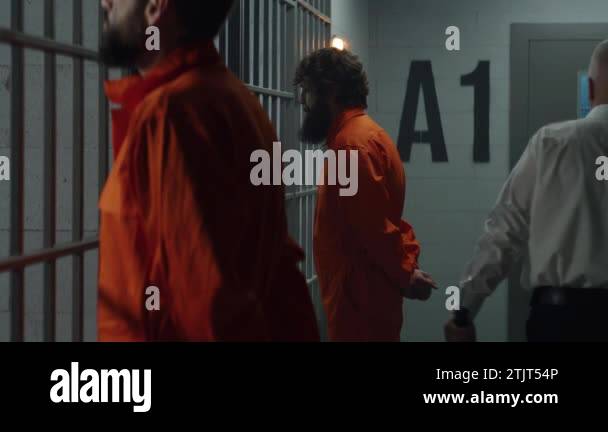 Two prisoners in orange uniforms stand facing the metal bars in front ...
