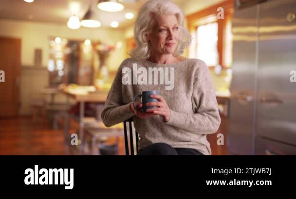 Lovely elder woman drinking coffee inside kitchen turning to smile at ...