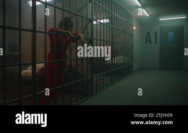 One female prisoner in orange uniform stands behind metal bars, another sits on the bed in ...