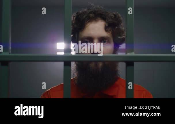 Portrait of male prisoner in orange uniform holding metal bars ...