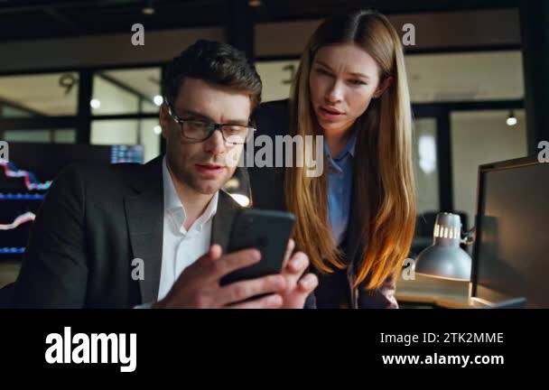 Two business people working dark finance office. Focused man woman using smartphone analysing ...