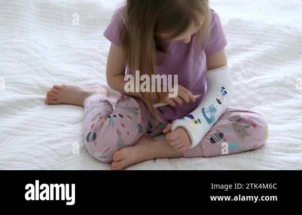 Adorable preschooler girl with a broken arm at home on the bed draws ...