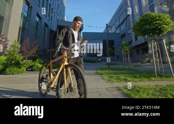 Side view of male with beard walking with bicycle, using smartphone ...