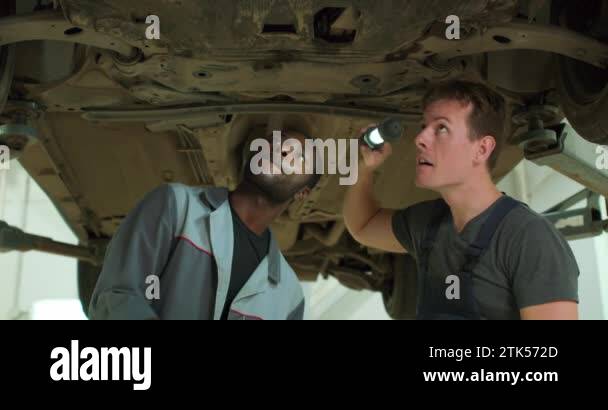 Two young hardworking auto mechanics working on undercarriage of car ...