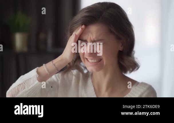 A young woman winces in pain, holding her head with both hands ...