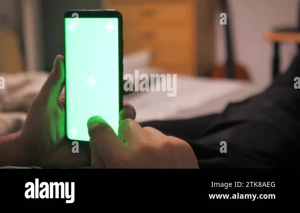 Man lying on couch using smartphone with chroma key green screen at night, scrdoing various ...