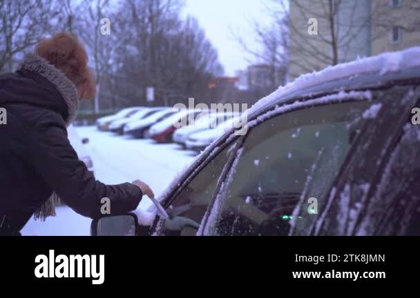 A woman clears snow from the side window of her car. The charms of the winter season when there ...