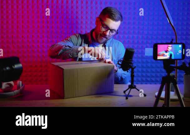 Young smiling blogger unpacking box with present sitting at home studio ...