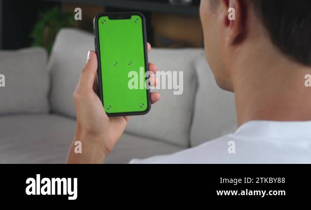 Male hold mobile phone in hand with chroma key green screen at living room. Man sitting at home ...