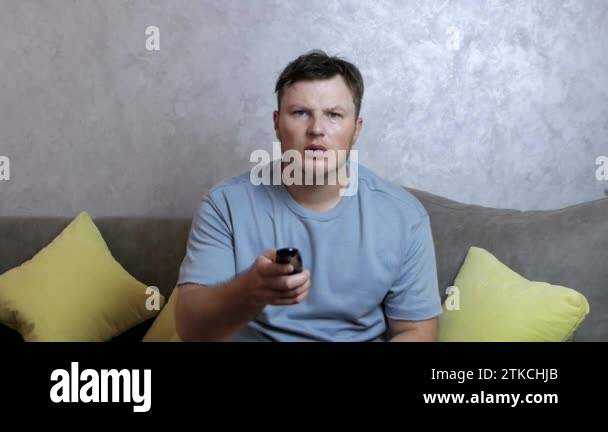 A young man is watching TV at home with a serious expression on his ...
