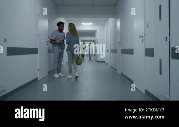 Woman goes medical center corridor, talks to African American doctor. Nurse and elderly patient ...