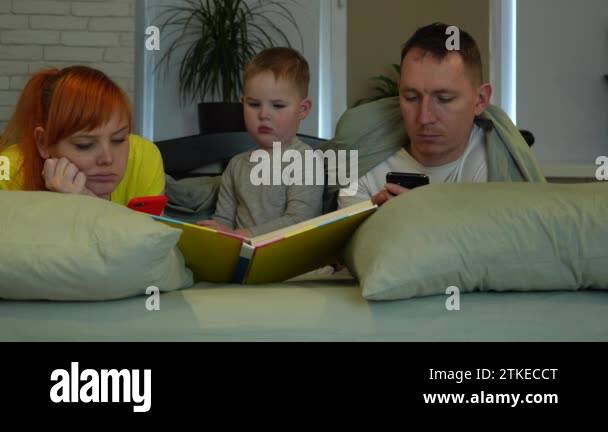 Young parents and little boy in bed. Mom and dad look at the screens of ...