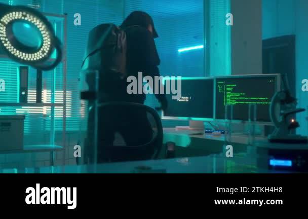 A hacker in a black hoodie and gloves is spying in the laboratory, typing on a computer keyboard ...