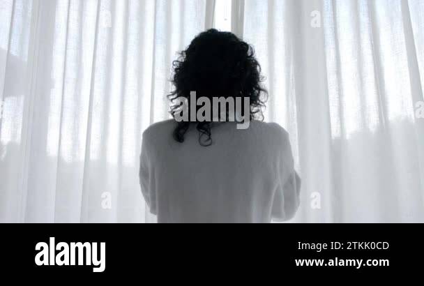 Biracial woman wearing bathrobe opening curtains and looking through ... Biracial woman wearing bathrobe opening curtains and looking through ...