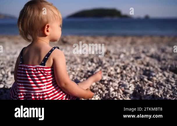 Little girl is sitting on a pebble beach and sorting through the ...