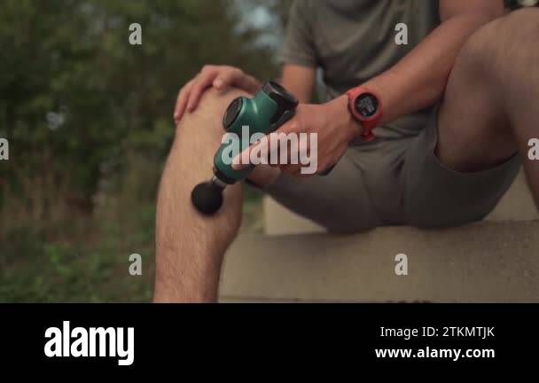 Young male athlete using a massage gun sitting on the outdoor sports ...