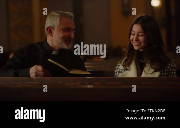 A priest sits in a church with a bible in his hands talking to a ...