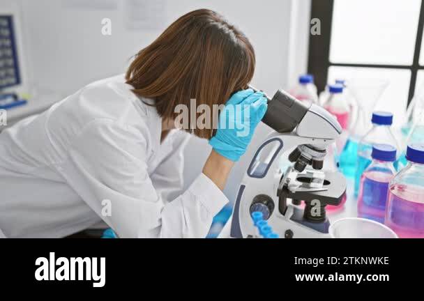 Young beautiful hispanic woman scientist using microscope sitting with arms crossed gesture at ...