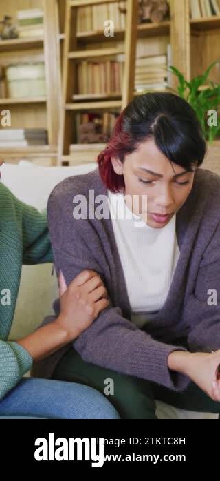 Vertical video of biracial sisters sitting on sofa and embracing, in ...
