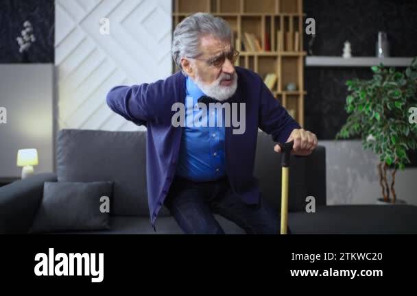 Senior man in glasses sits with a cane, got up from the sofa, his back ...