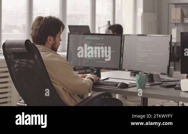Rear view slowmo of male programmer typing sitting in front of two computer monitors in office ...