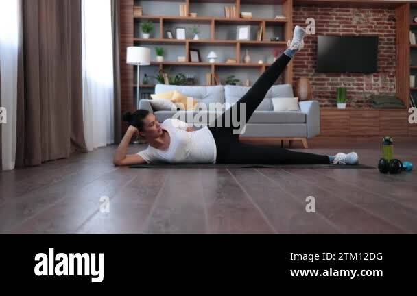 Multiethnic charming woman dressed in sport clothes lying on yoga mat at living room and lifting ...