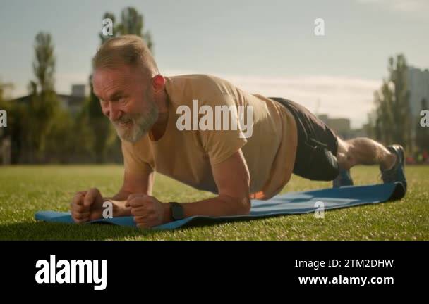 Older man standing plank pose on lawn stadium city outdoors physical exercise endurance strength ...