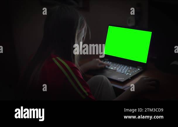 School-age girl sits alone in dark room in front of laptop screen and ...