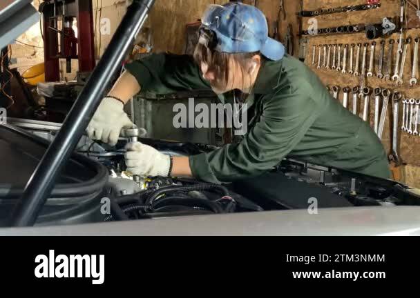 A young woman in coveralls works at a car service station performs ...