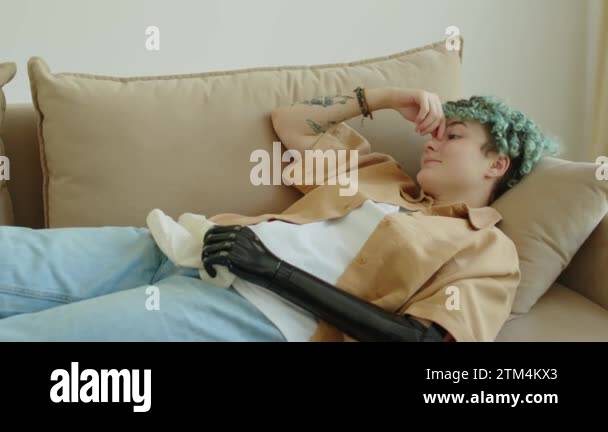 Sad gen Z girl with prosthetic arm and blue hair lying on sofa, holding ...