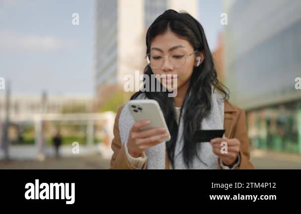 Young Asian Customer Holding Credit Card and Smartphone Wearing Earphones. Lady Shopper Using ...
