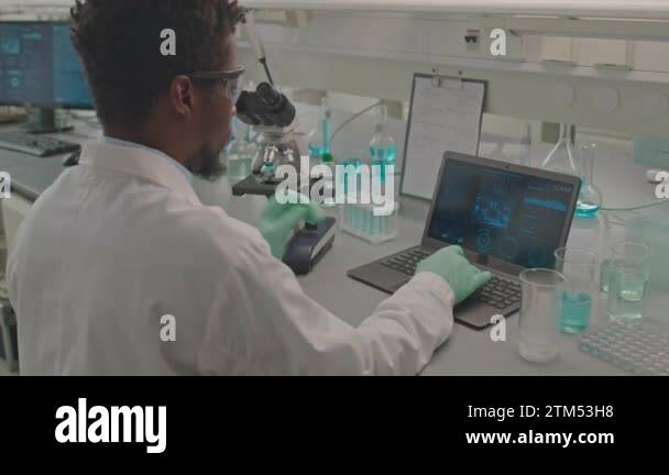 Waist up of young African American male scientist in white lab coat looking through microscope ...