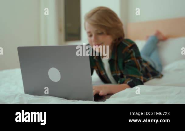 Teenage child boy using laptop. Child typing on the keyboard lying in bed. Play video game ...