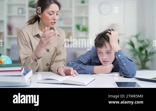 A tired mother is doing homework with her school boy child scolding her son while helping with ...