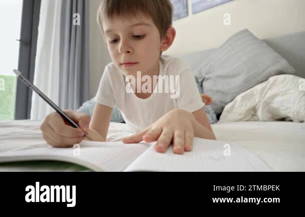 Young boy in bed, writing in a notebook as he does his homework ...