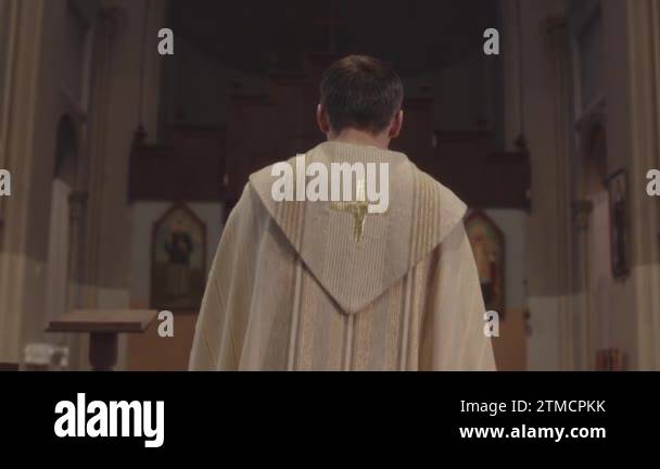 Tracking rear view slowmo of pastor in white alb walking along Catholic ...