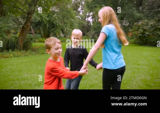Three young children playing ring around the rosie in a park in slowmo ...