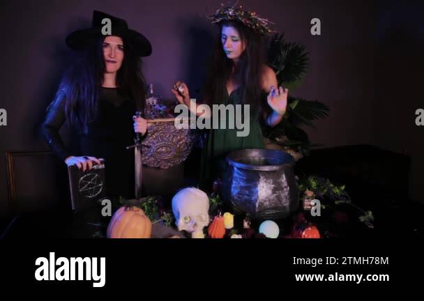 Two witches pose standing next to a table with a cauldron, skull and ...