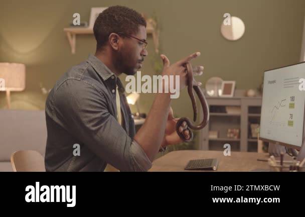 Medium side view slowmo of young Black hipster man in eyeglasses and denim shirt sitting at desk ...