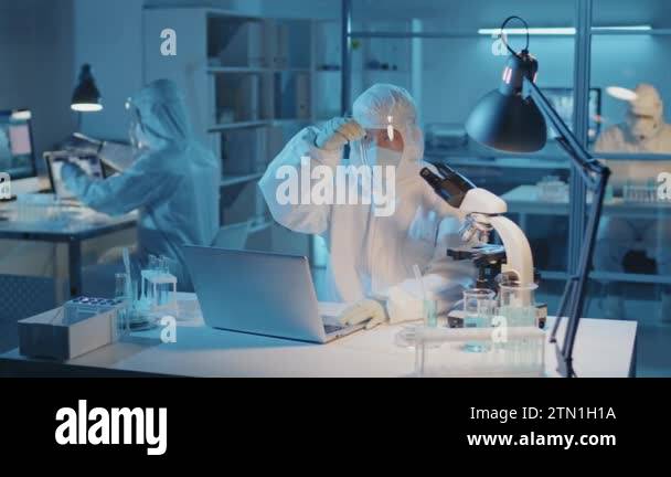 Waist-up of Asian scientist in white hazmat suit, respirator and face shield sitting at desk in ...